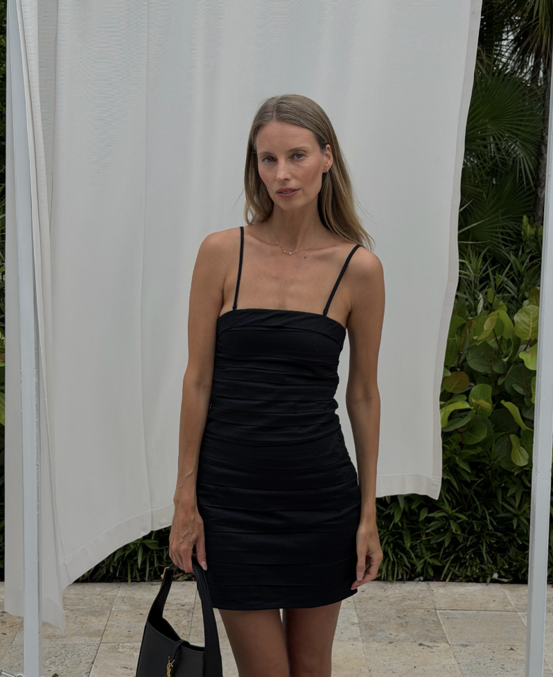 Woman in a black dress stands under a white cabana%2C holding a black purse. The background is lush with green foliage. The mood is elegant and calm.